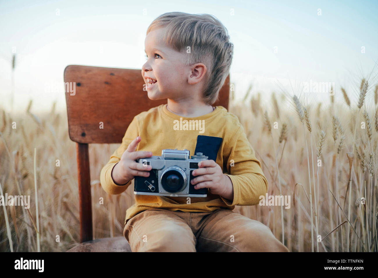 Cheerful boy with camera looking away while sitting on chair amidst ...