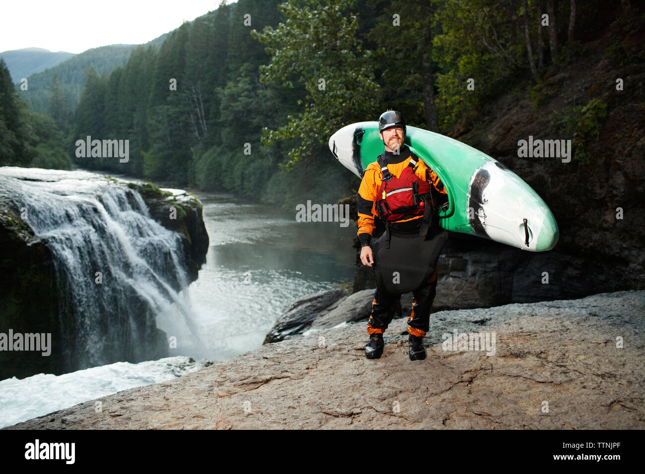 Male kayaker standing on rock with waterfall and trees in background ...
