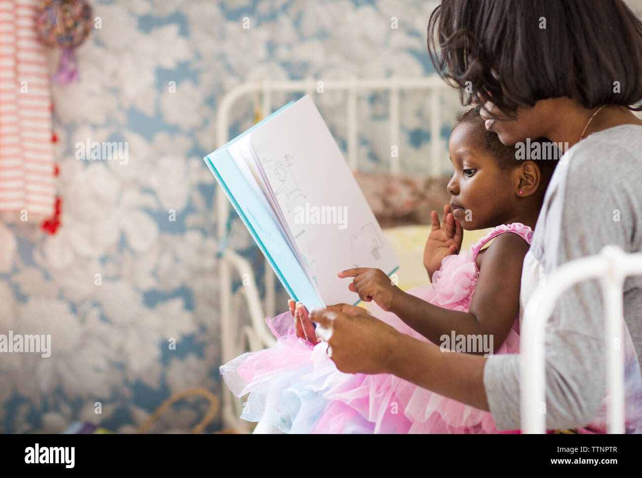Girl pointing at picture book while sitting with mother in bedroom ...