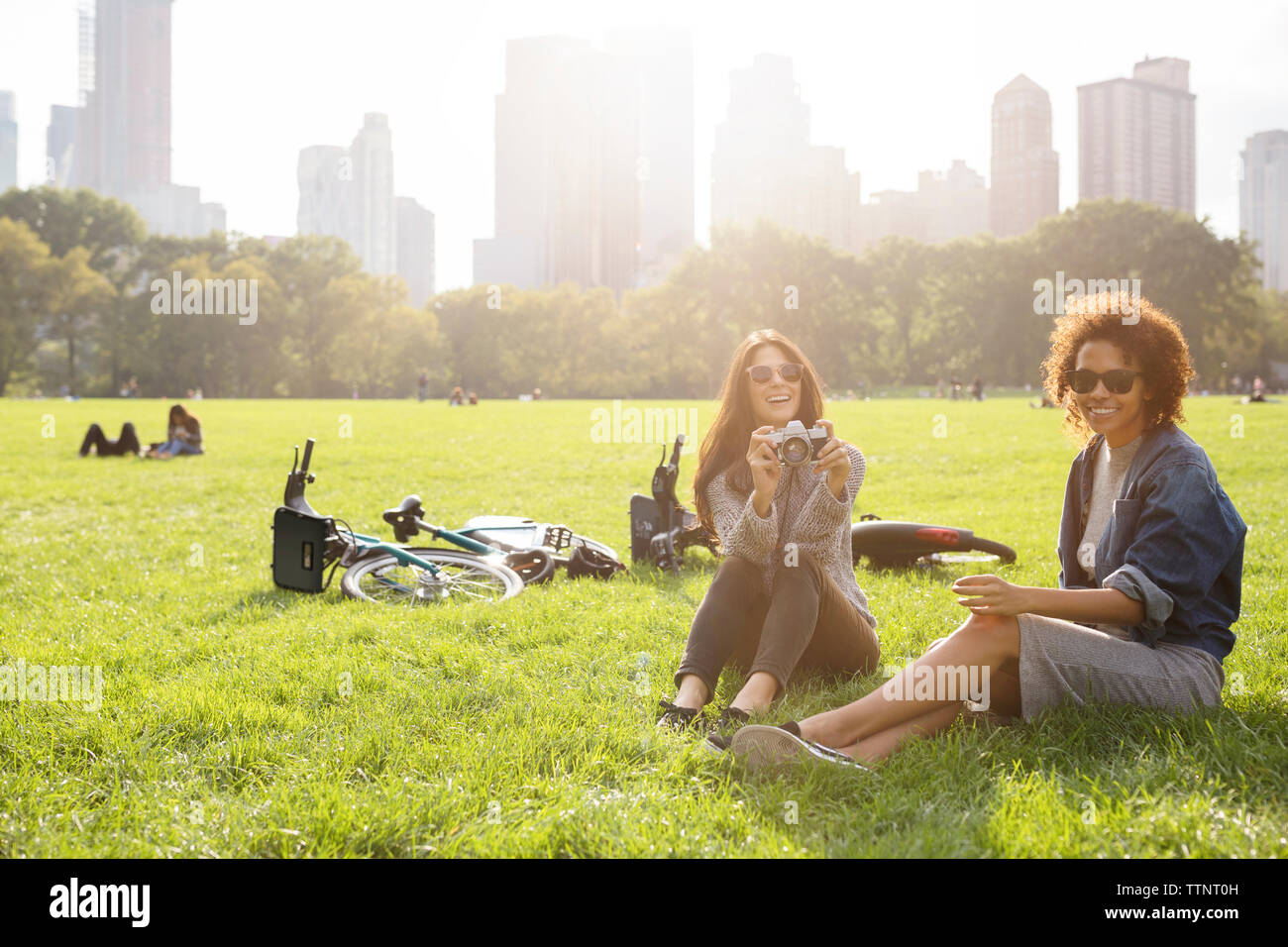 Happy woman holding camera while relaxing with friend on grassy field ...