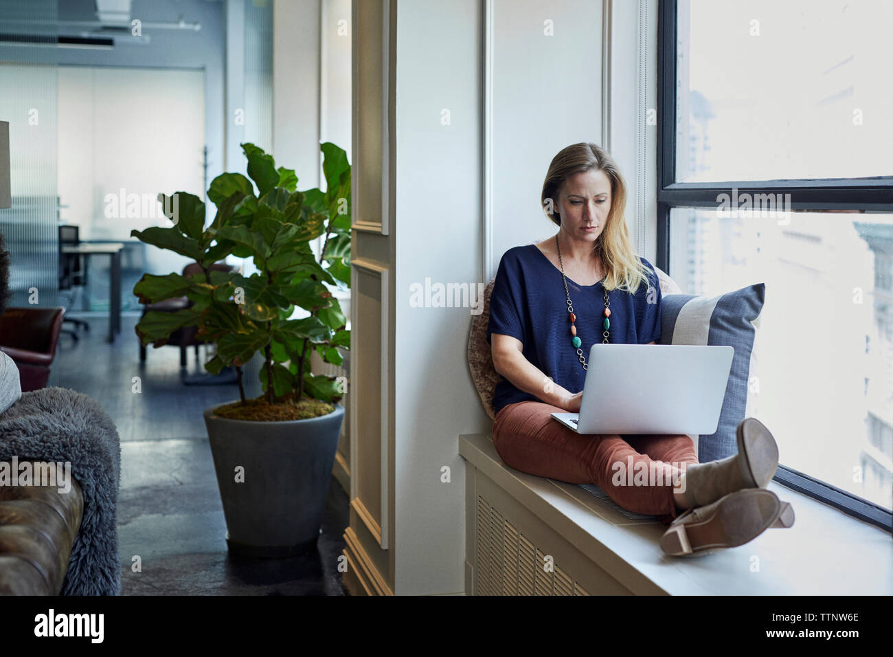 businesswoman using laptop computer while sitting on window sill in ...