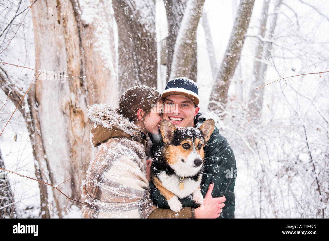 Couple huddled together laughing with dog in forest during snowfall ...