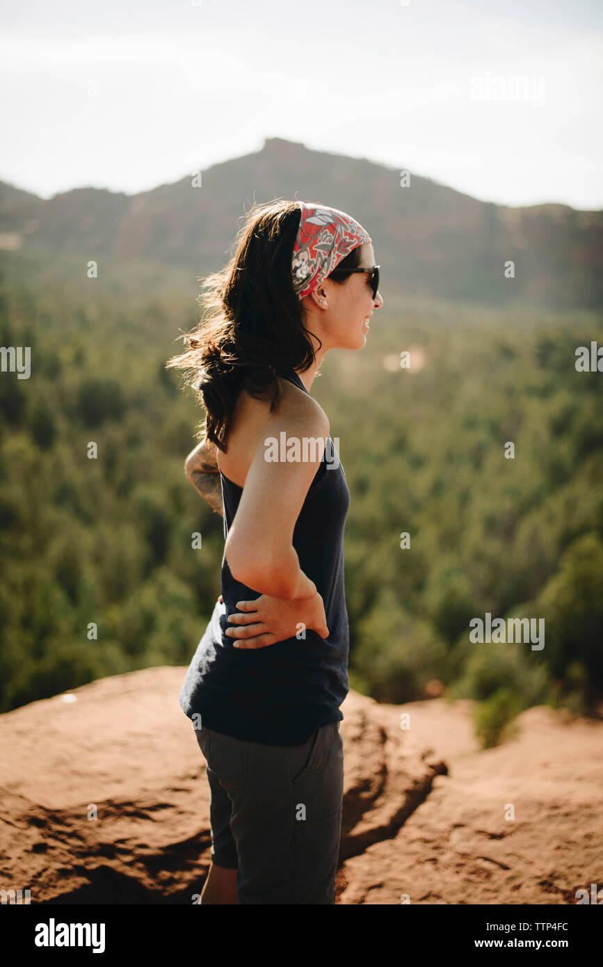 Side view of hiker standing with hand on hip at cliff against forest ...