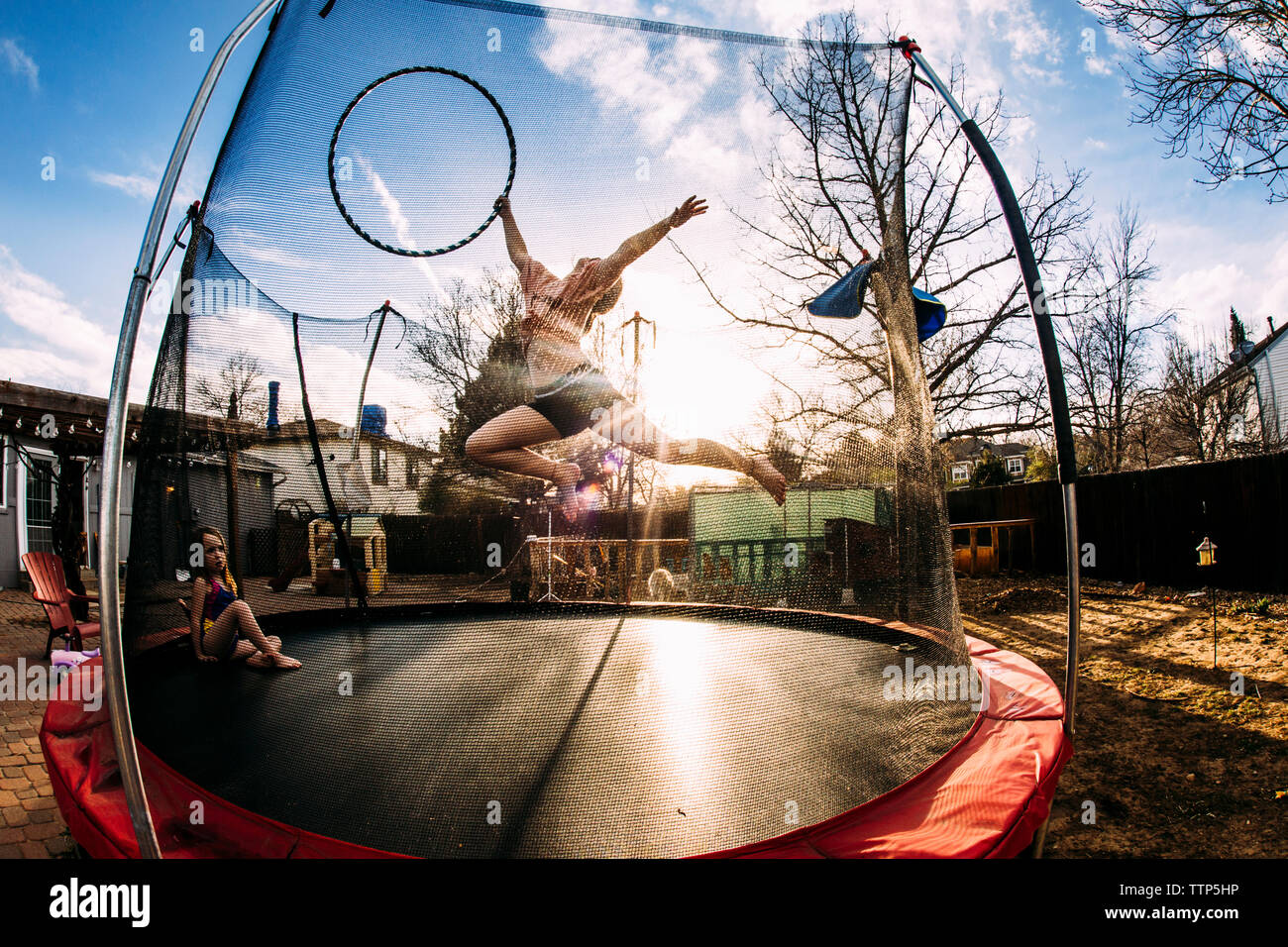 Girl with plastic hoop jumping while sister sitting in trampoline at ...