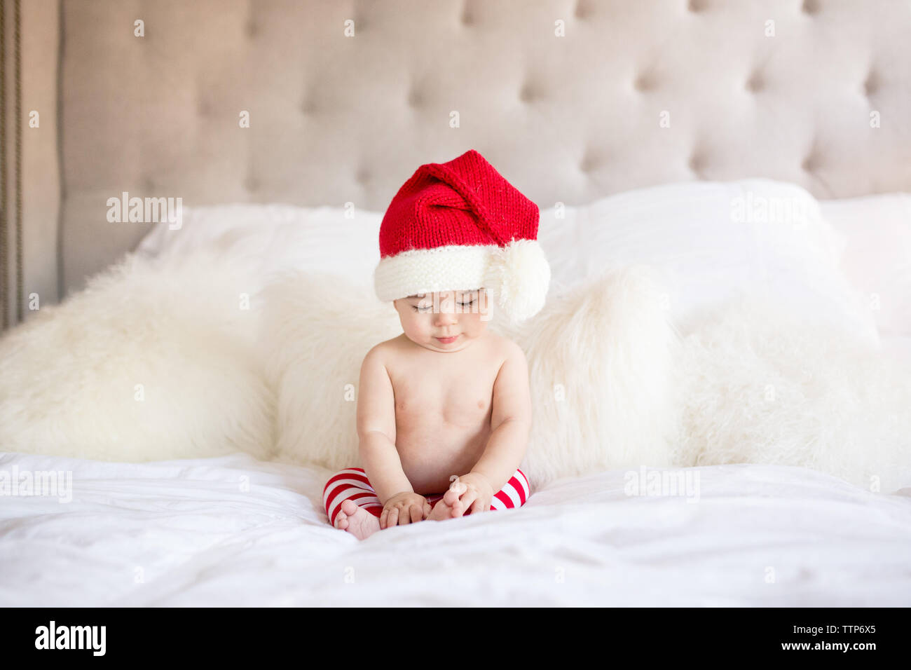 Cute baby boy sitting on bed inside with Santa hat on looking at feet ...