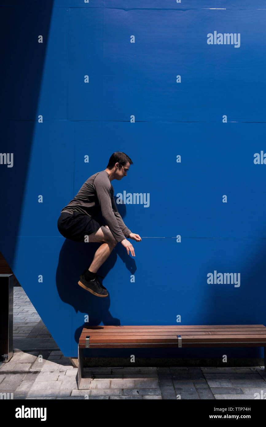 Side view of man jumping on wooden bench against blue wall during sunny ...