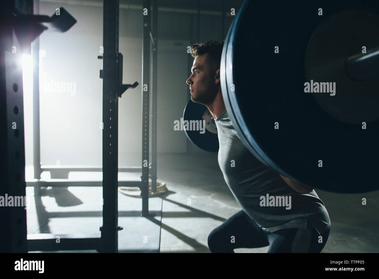 Side view of male athlete looking away while lifting barbell at gym ...