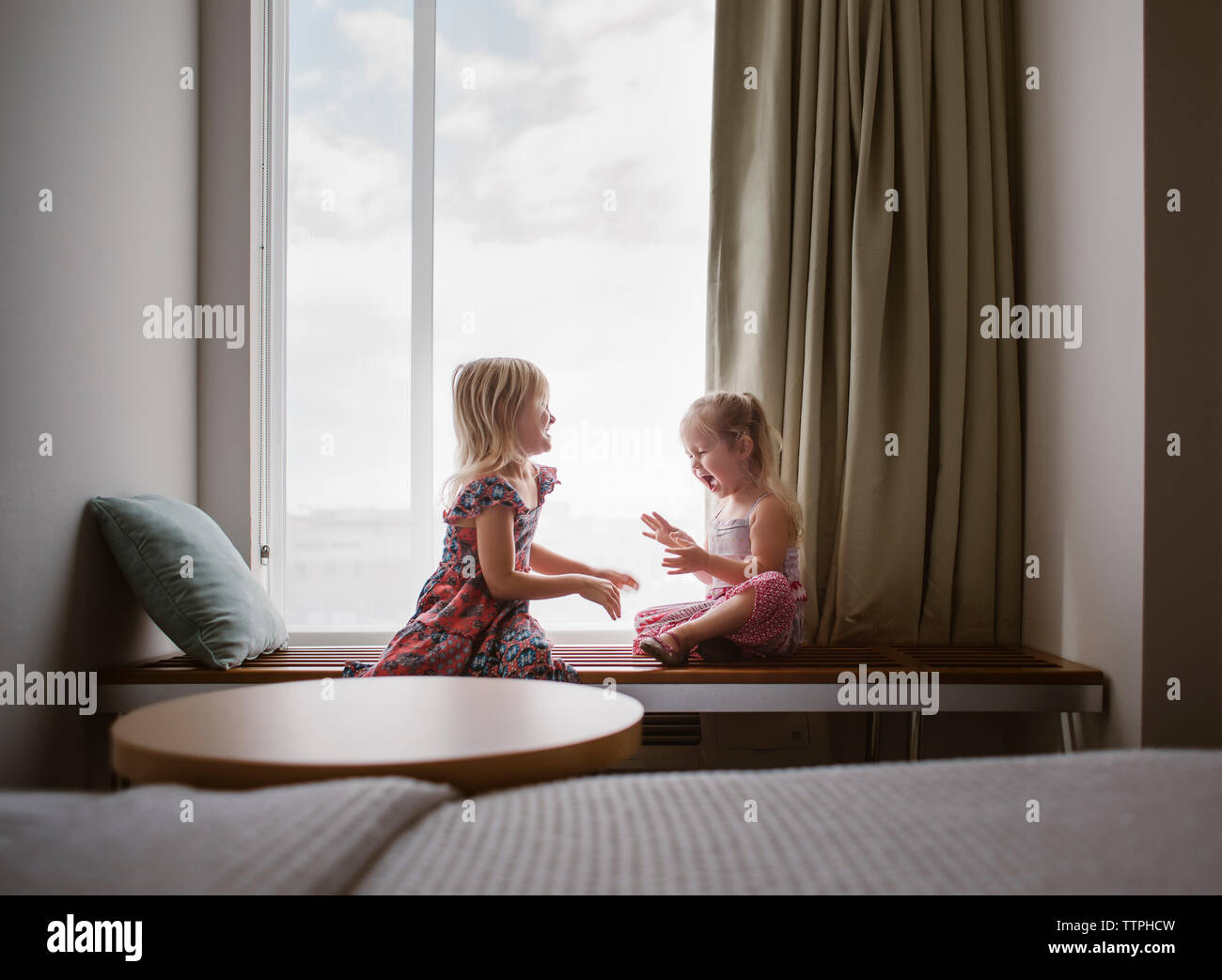 Happy sisters talking while sitting on table by window at home Stock ...