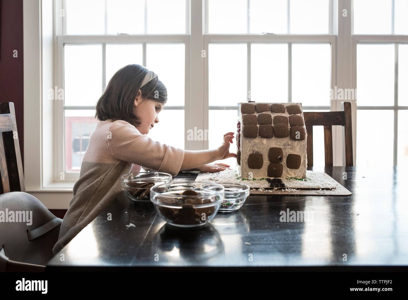 Side view of girl making gingerbread house on table against window at ...