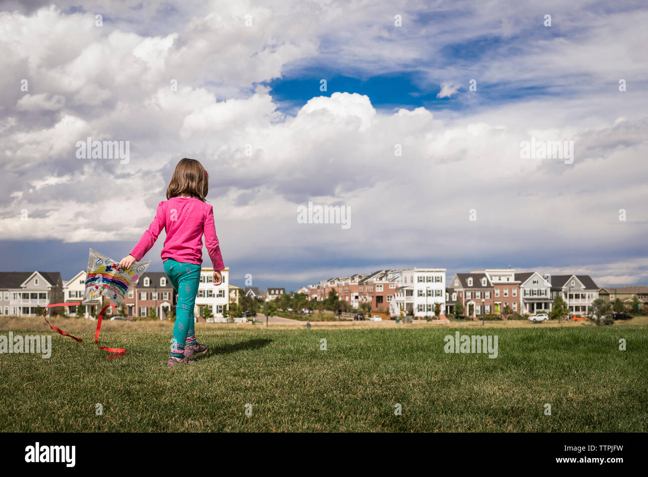Rear view of girl with kite walking on grassy field against cityscape ...