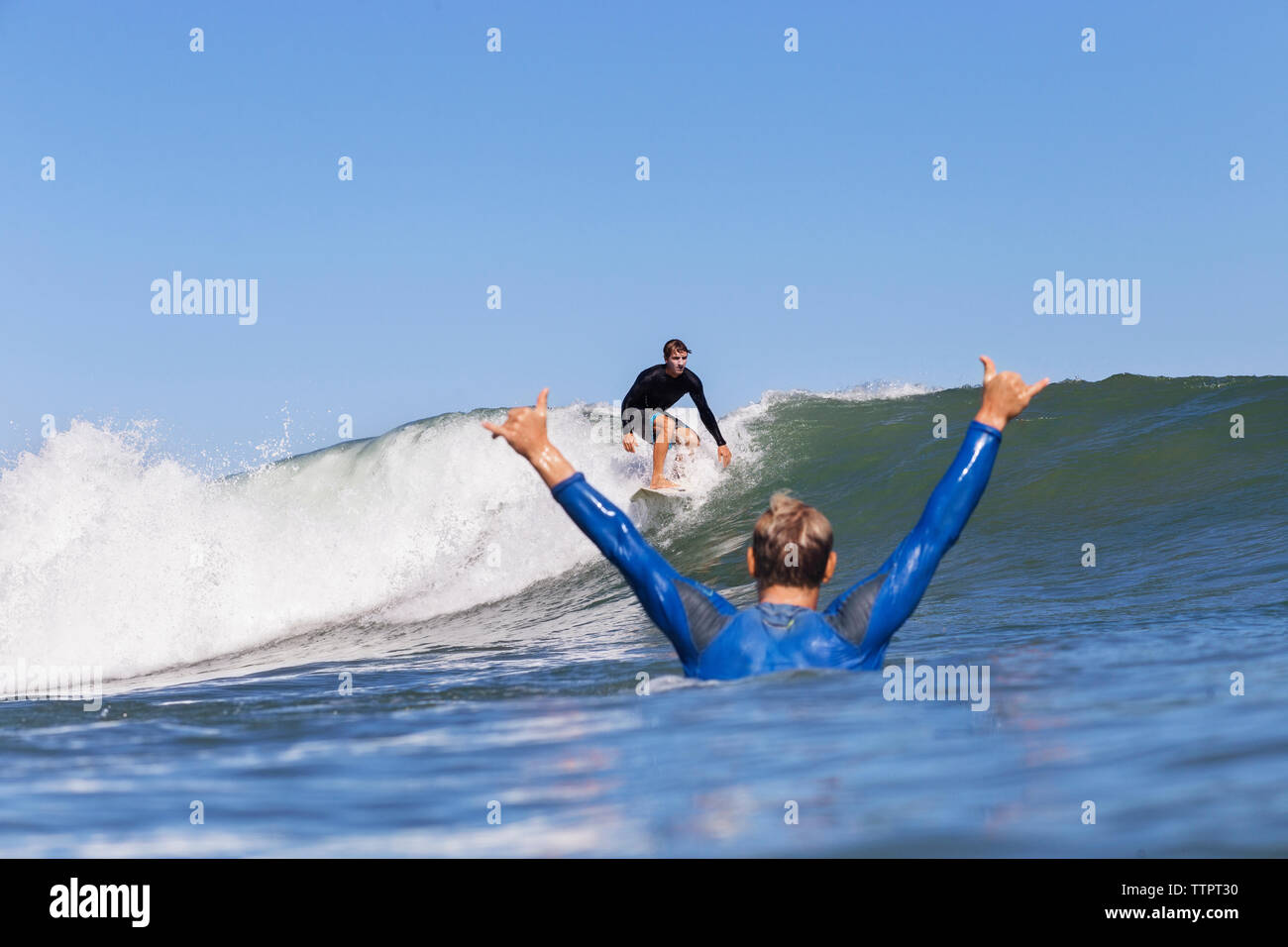 Man cheering while looking at friend surfing on sea against clear blue ...