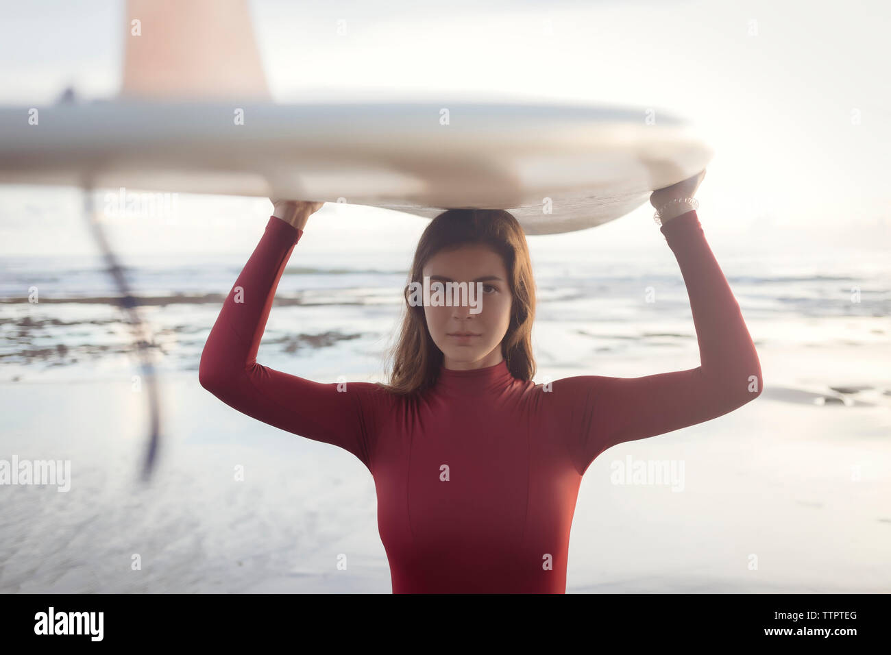 Portrait of young woman carrying surfboard on head while standing at ...
