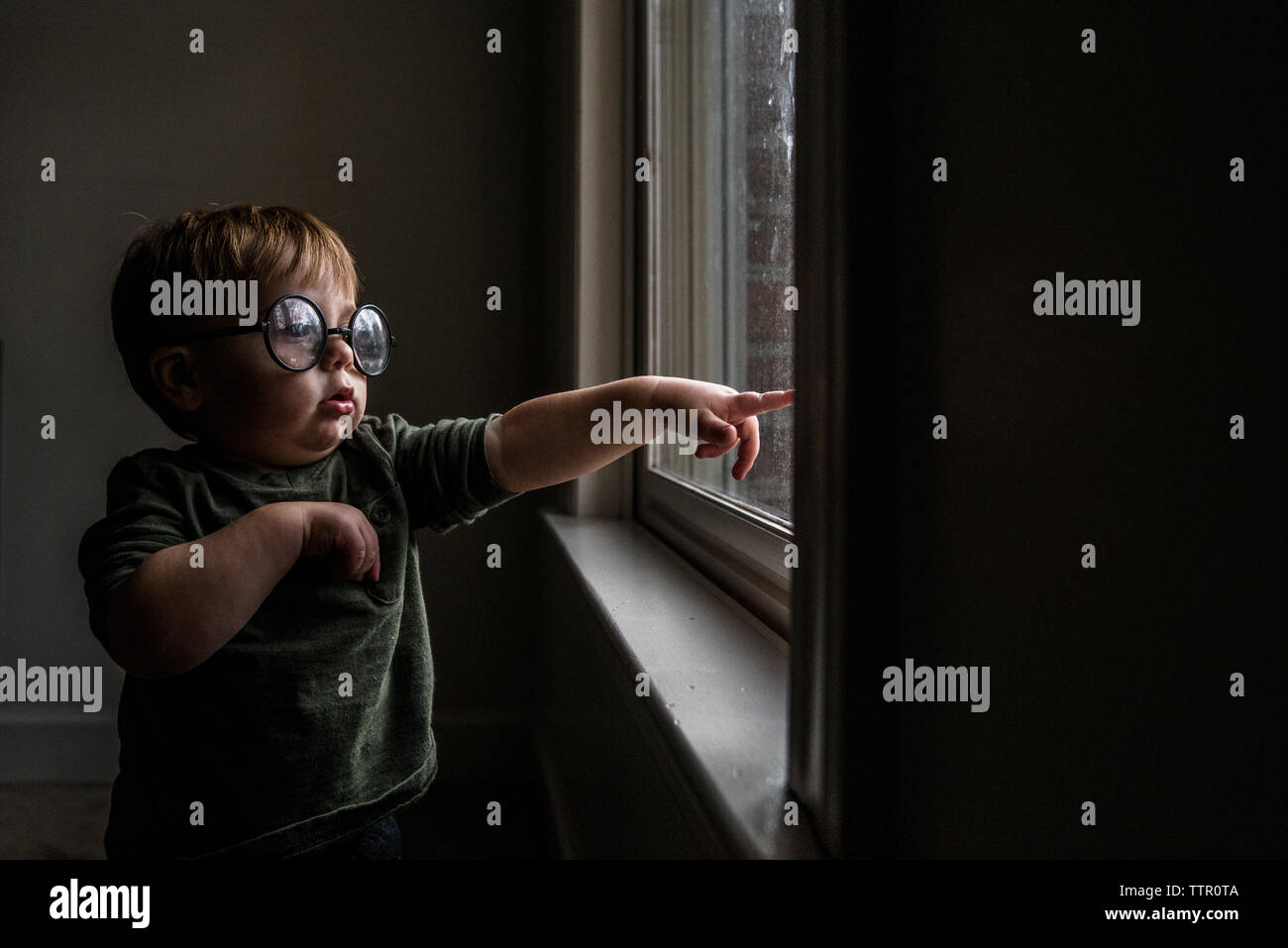 Toddler boy wearing round glasses standing by window pointing outside ...