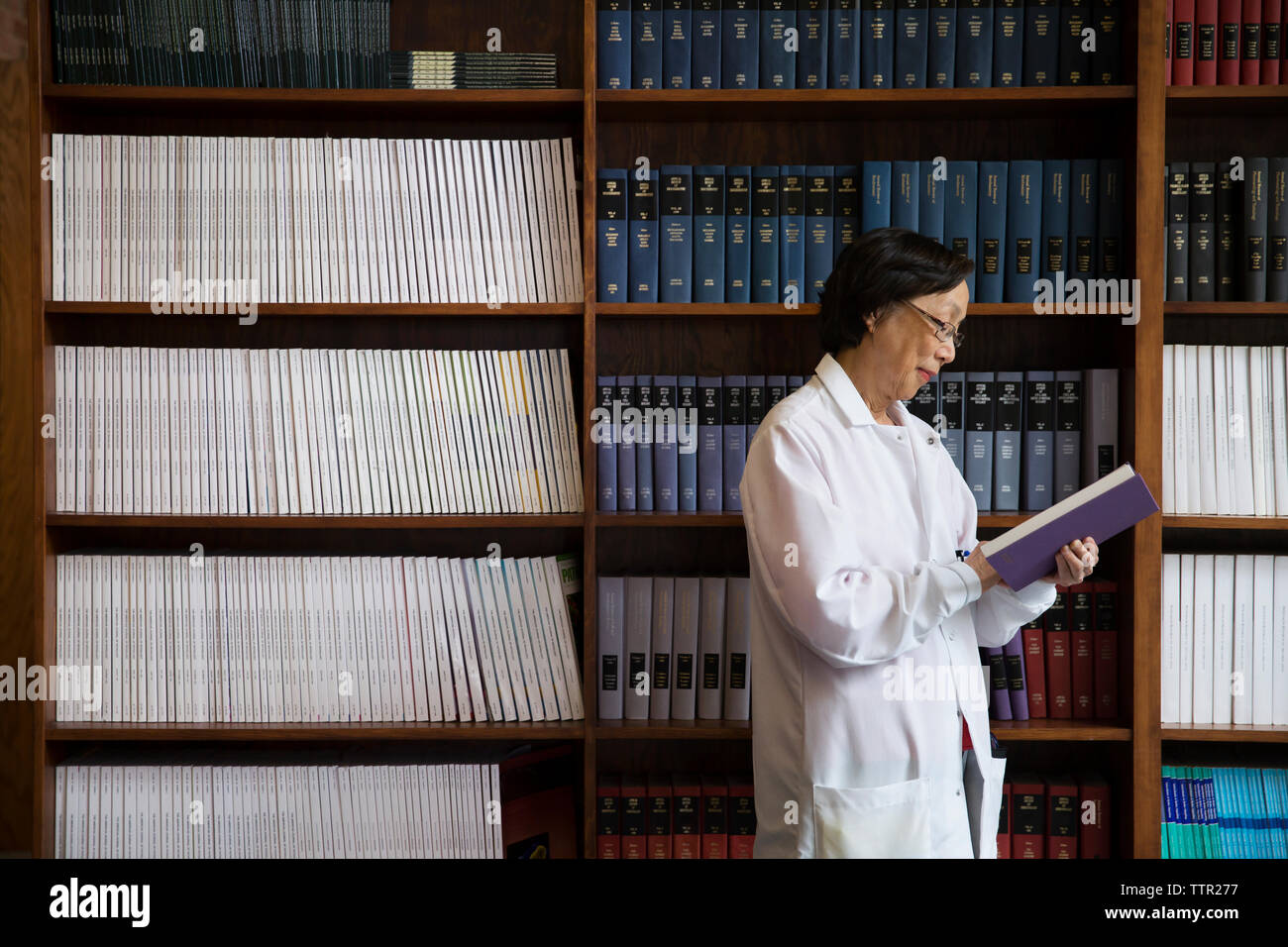 Side view of senior female scientist reading book by shelves at ...