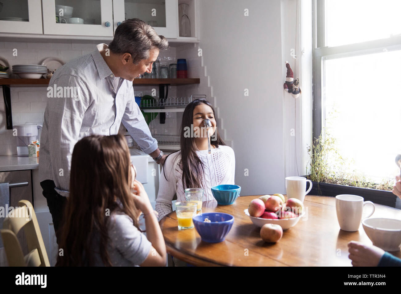 Father looking at girl balancing spoon on nose at table in kitchen ...