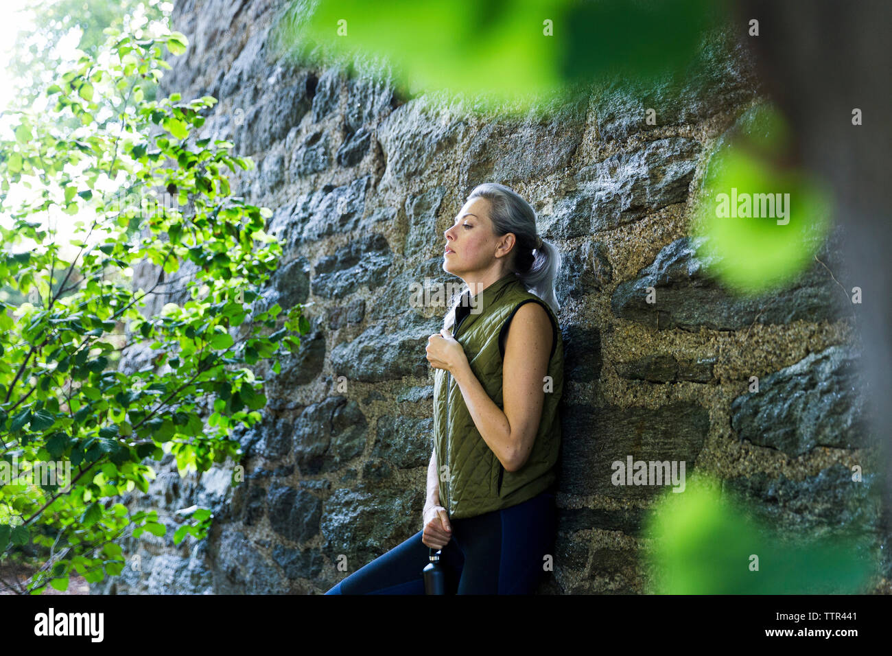 Woman closing zipper of jacket while leaning on stone wall Stock Photo ...