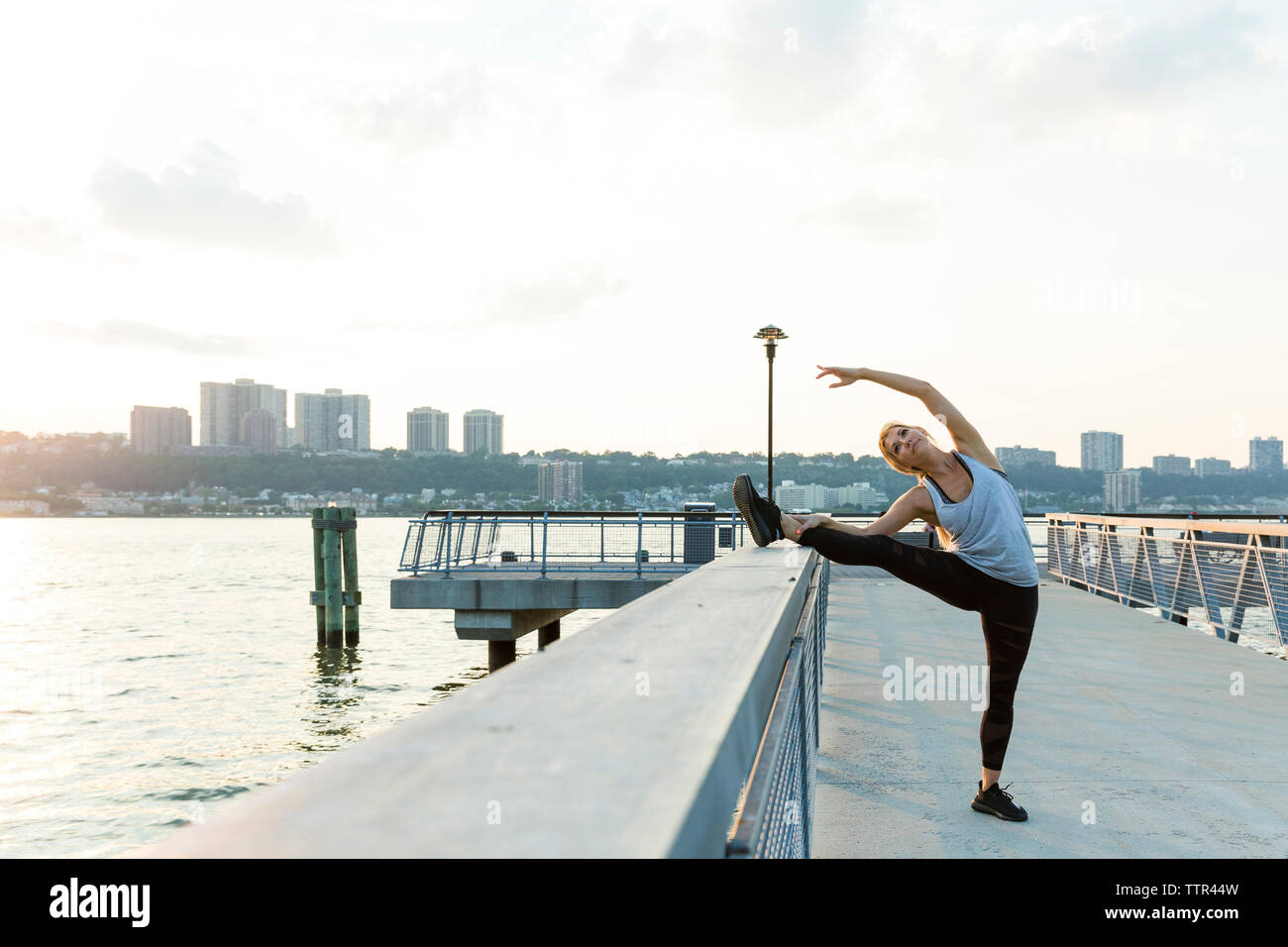 Full length of woman stretching leg on railing at bridge against sky in ...