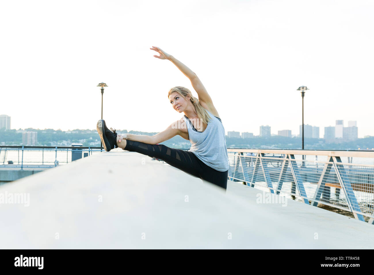 Woman stretching leg on railing at bridge against clear sky in city ...