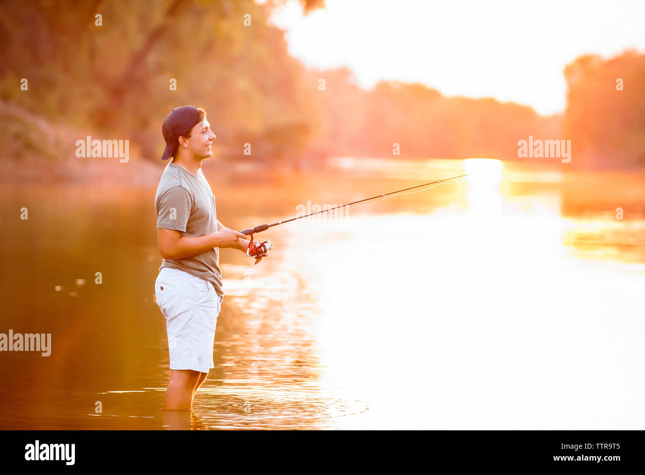 Side view of young man fishing while standing in lake during sunset ...