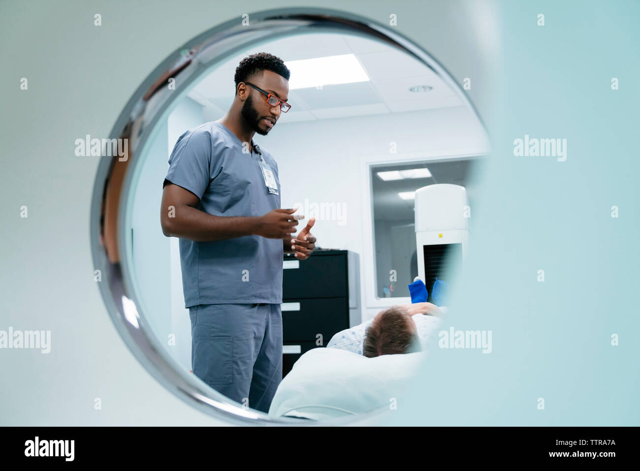 Male nurse talking with patient lying in examination room seen through ...