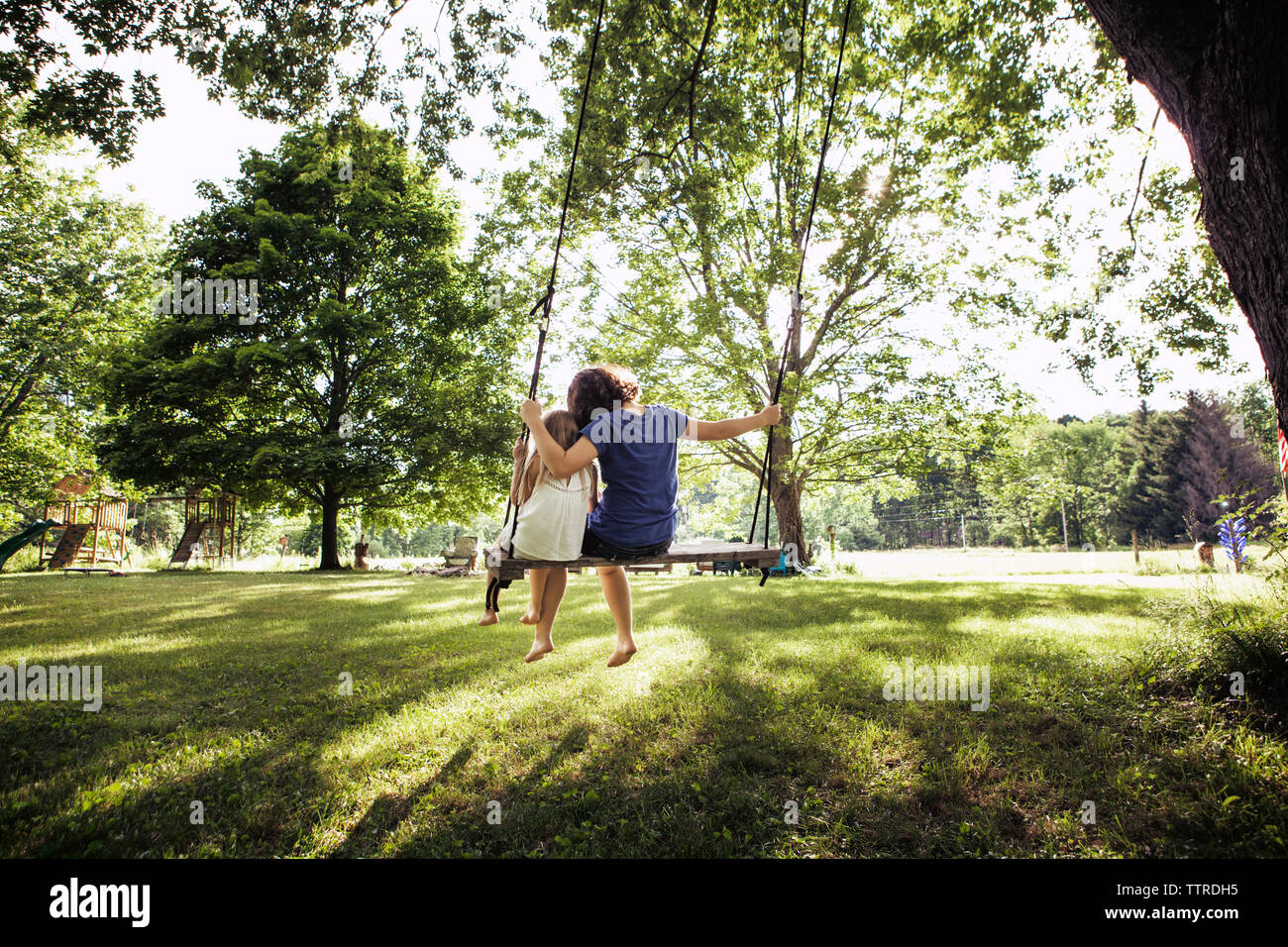 Rear view of sisters sitting on swing at grass field Stock Photo - Alamy
