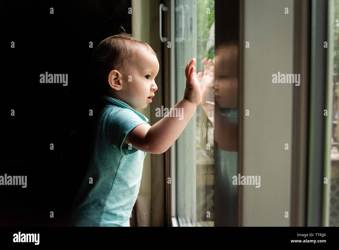 Side view of cute baby boy looking through window while standing in ...