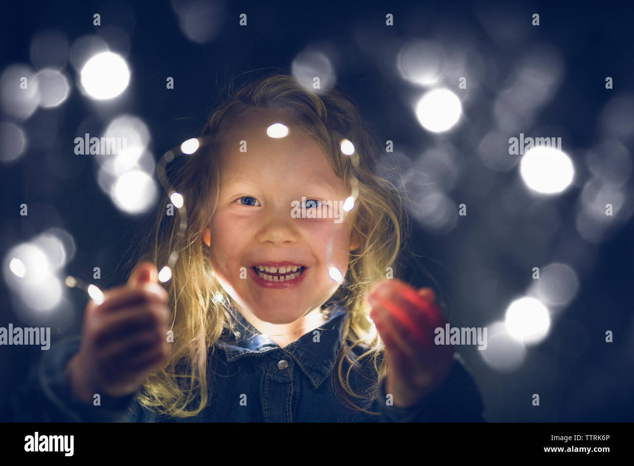 Portrait of cheerful girl playing with illuminated string lights during ...