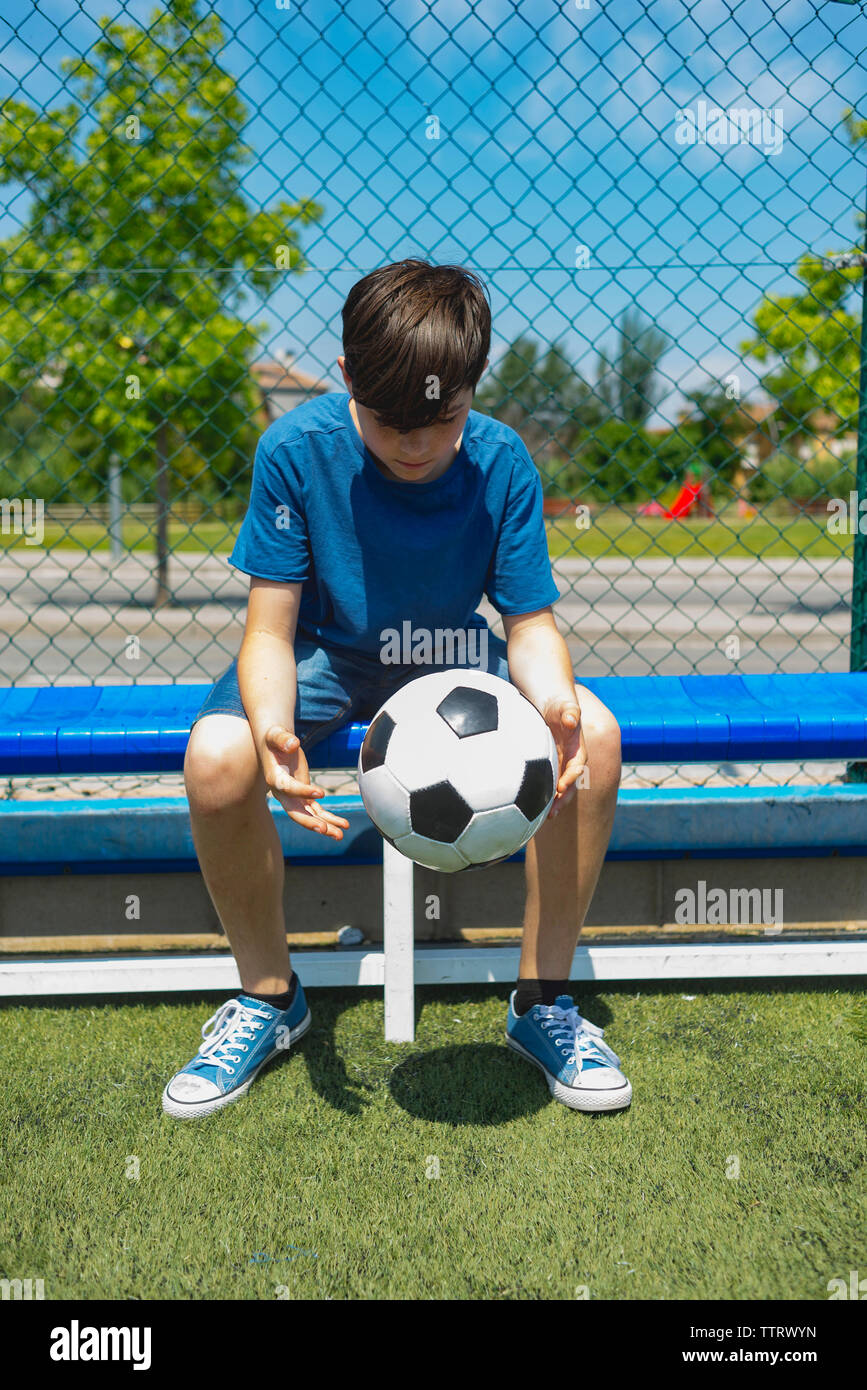 Boy holding soccer ball while sitting on bench by fence at dugout Stock ...