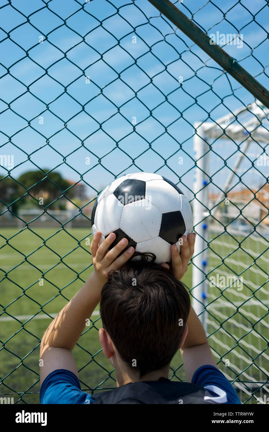 Rear view of boy holding soccer ball while standing by fence at dugout ...