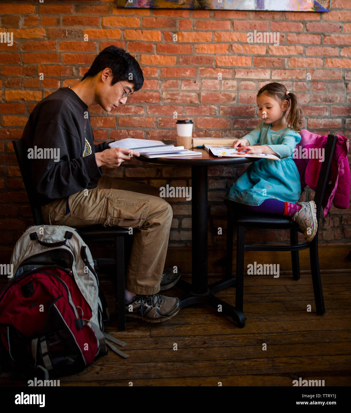 Father working while daughter reading book while sitting against brick ...