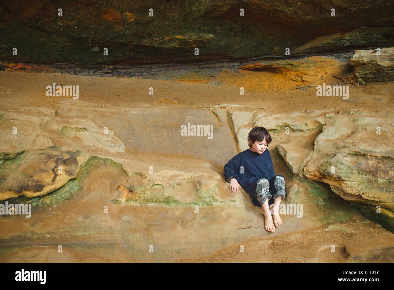 A small child sits barefoot cradled on rocky sandstone ledge in gorge ...