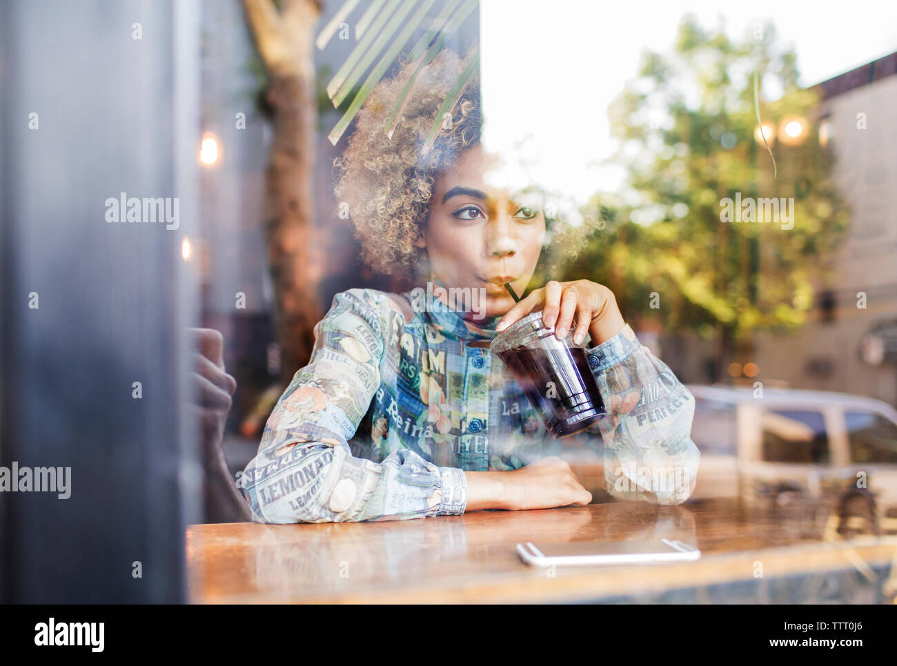 Woman drinking ice tea while sitting at cafe seen through glass window ...