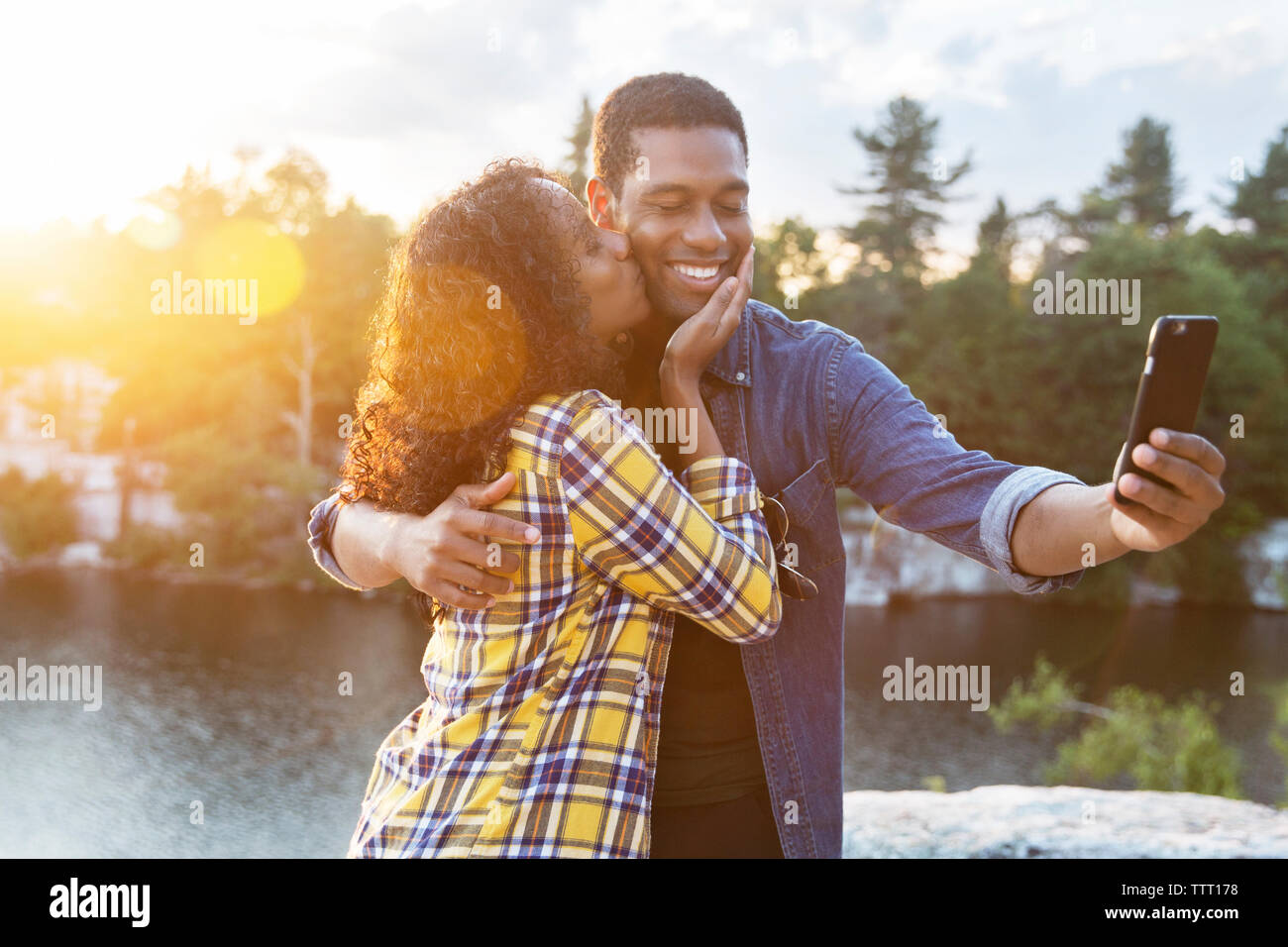 Woman kissing man taking selfie through smart phone by lake during ...