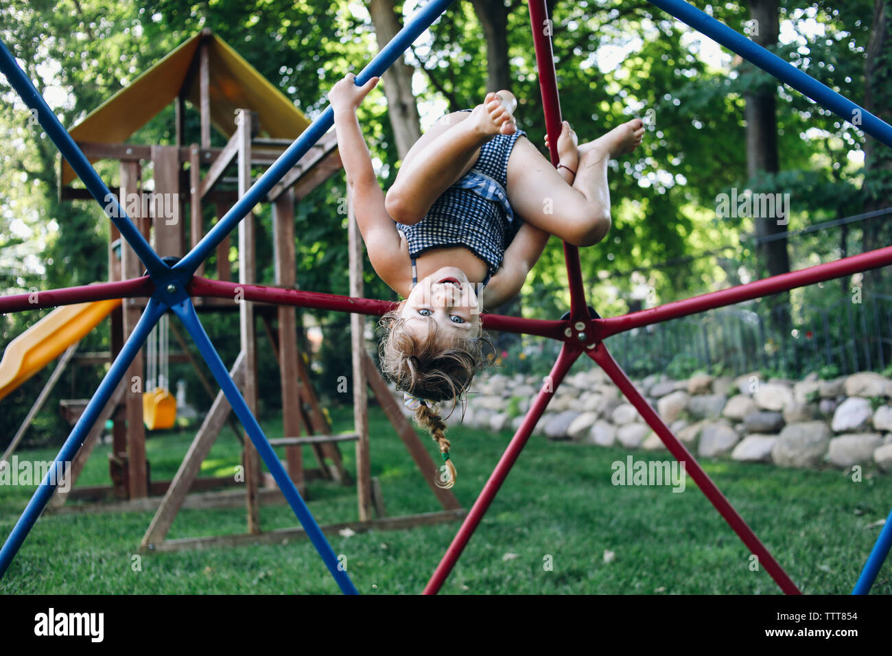 Portrait of cute smiling girl hanging upside down on jungle gym against trees at playground ...
