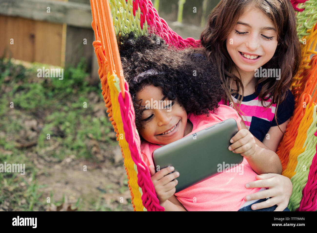 Happy Siblings Using Tablet While Sitting On Hammock At Backyard Stock