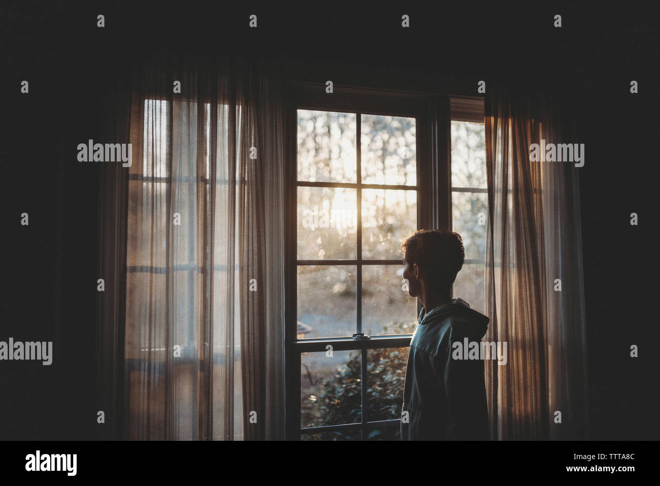 Side view of teenage boy looking through window while standing at home ...