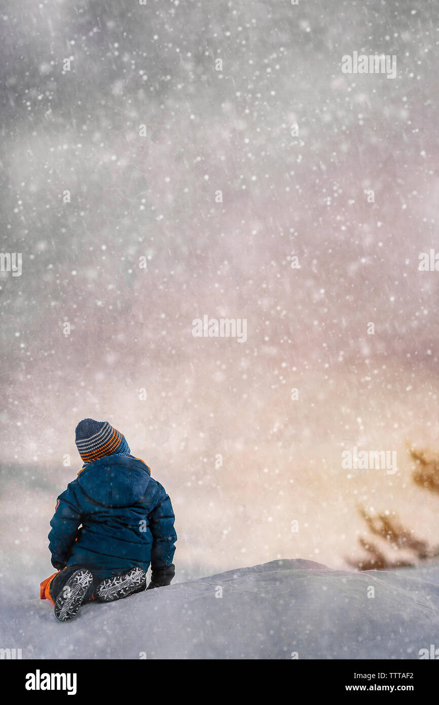 Rear view of boy sitting on snow covered field during snowfall at ...