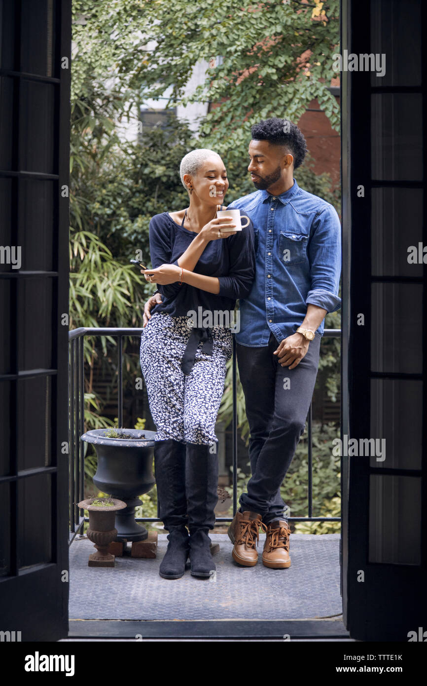Happy couple talking while standing in balcony seen through doorway ...