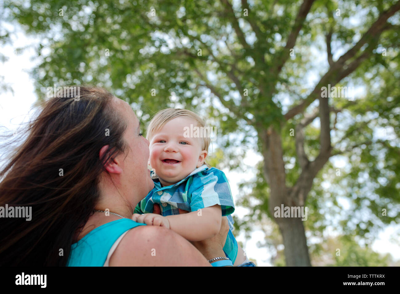 Low angle portrait of cheerful son being carried by mother against tree ...