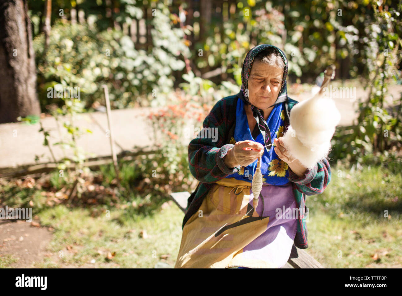 Senior woman spinning thread on spindle while sitting on bench at yard ...