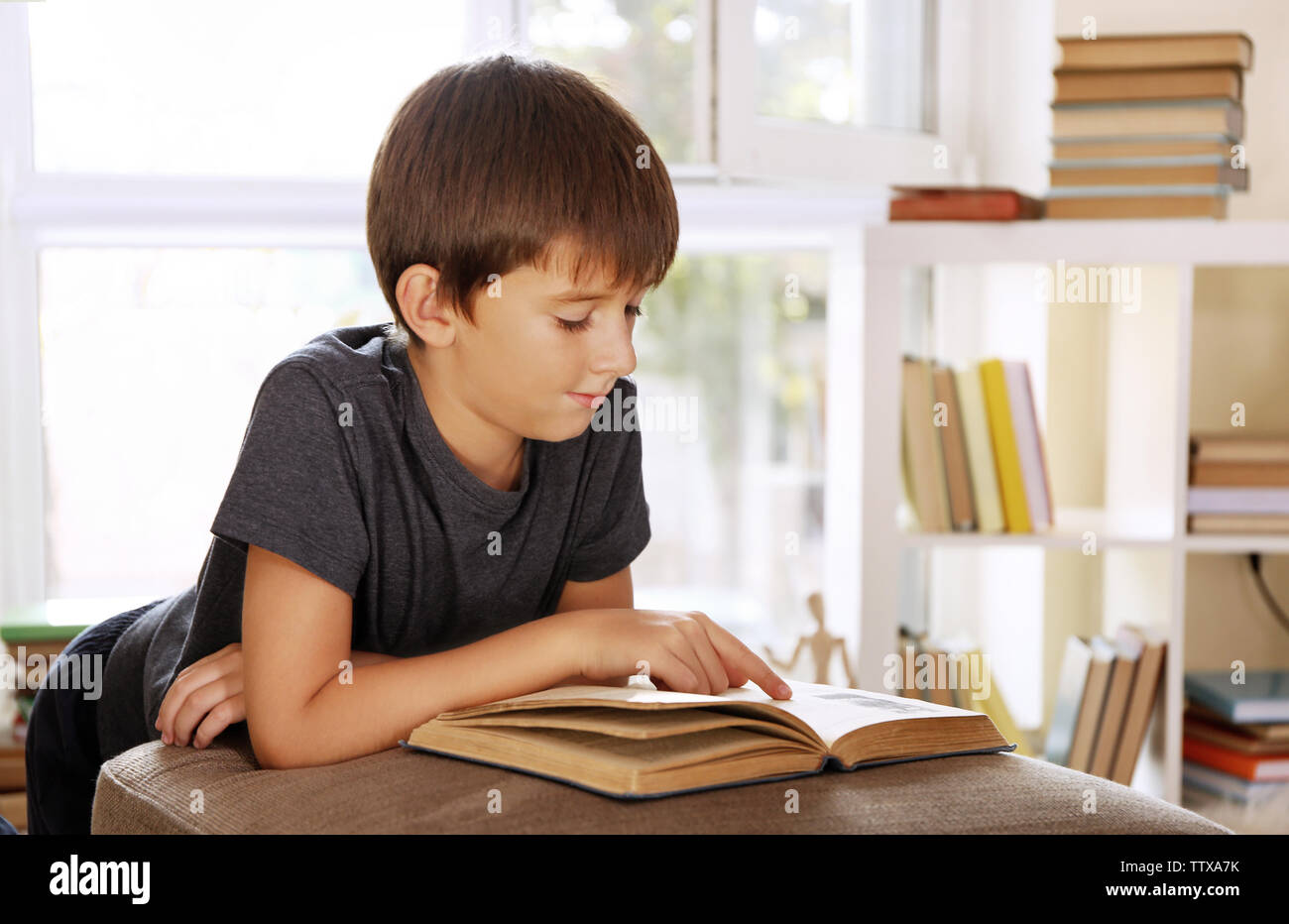 Cute schoolboy reading interesting book on pouf in room Stock Photo - Alamy