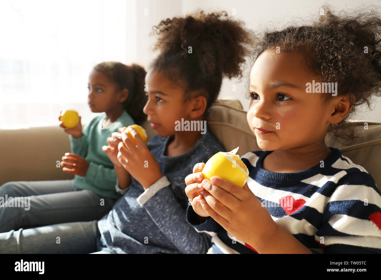 Three cute African girls sitting on sofa and eating lemons Stock Photo ...