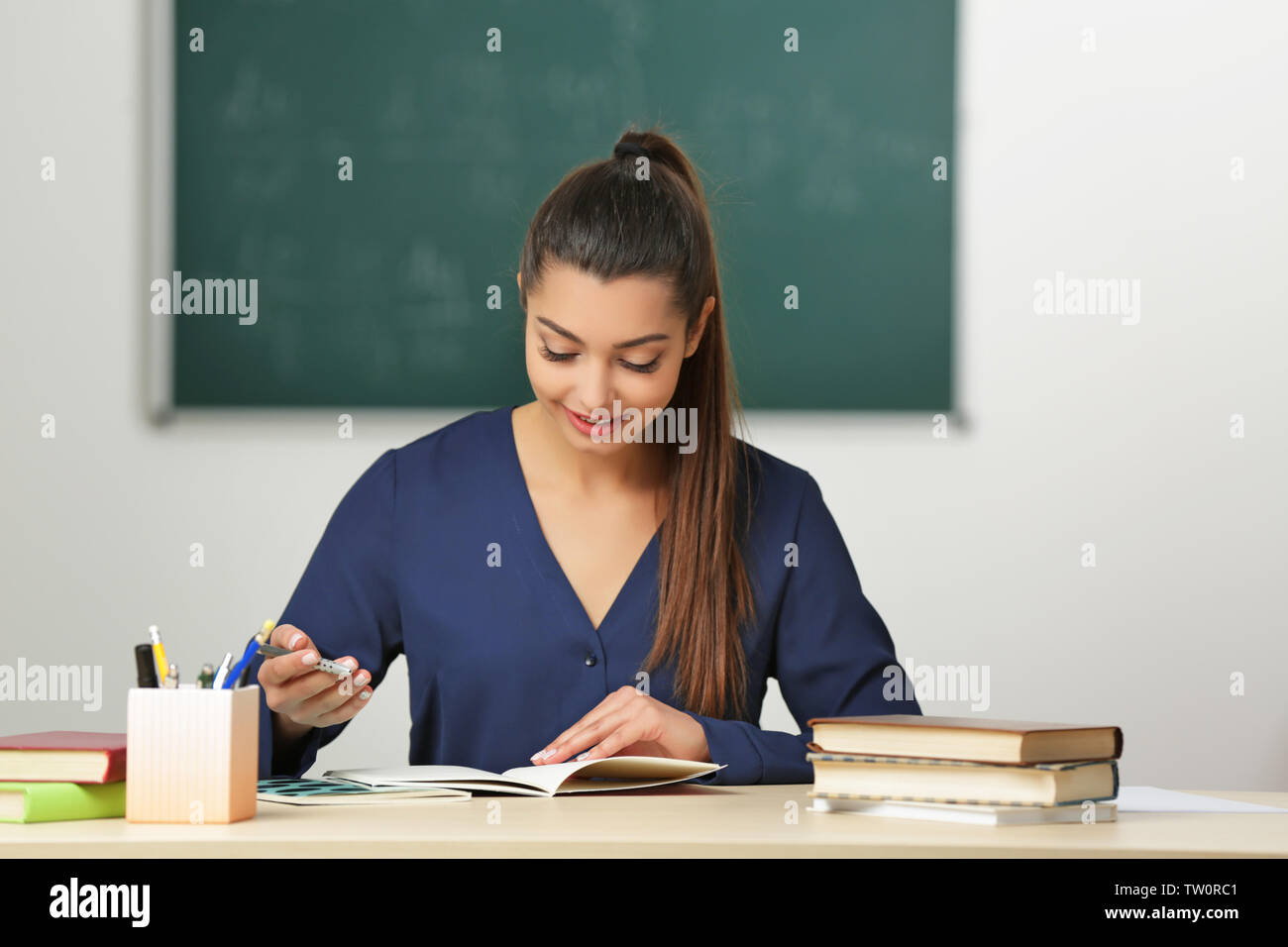 Beautiful young teacher sitting at table in classroom Stock Photo - Alamy
