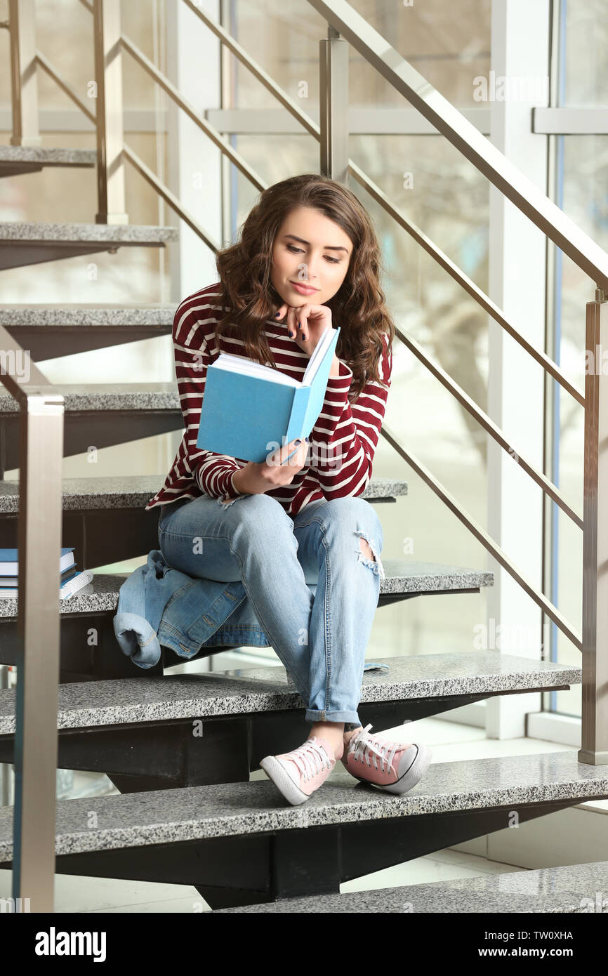 Beautiful young woman reading book while sitting on stairs near window ...