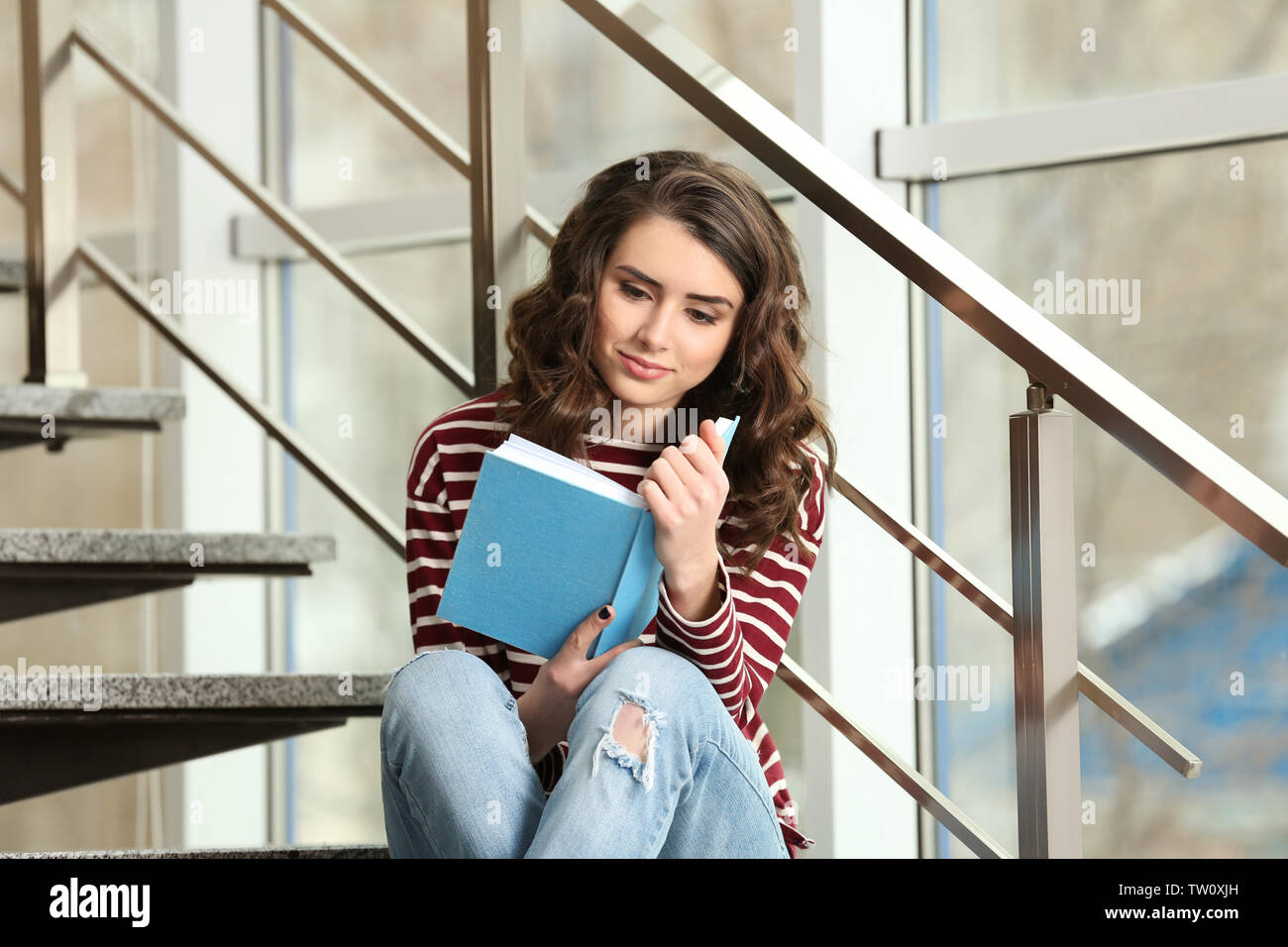 Beautiful young woman reading book while sitting on stairs near window ...