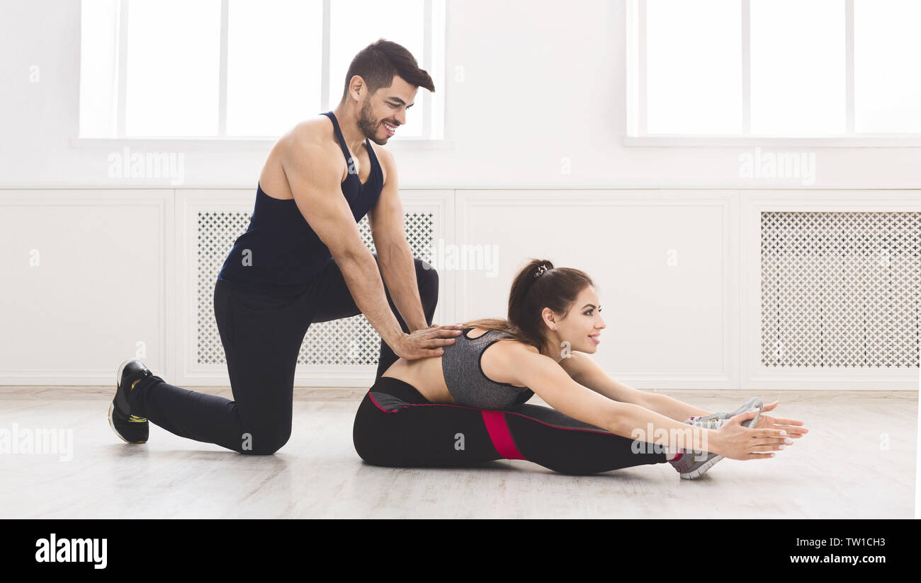 Young muscular coach helping girl doing stretching exercise in light ...
