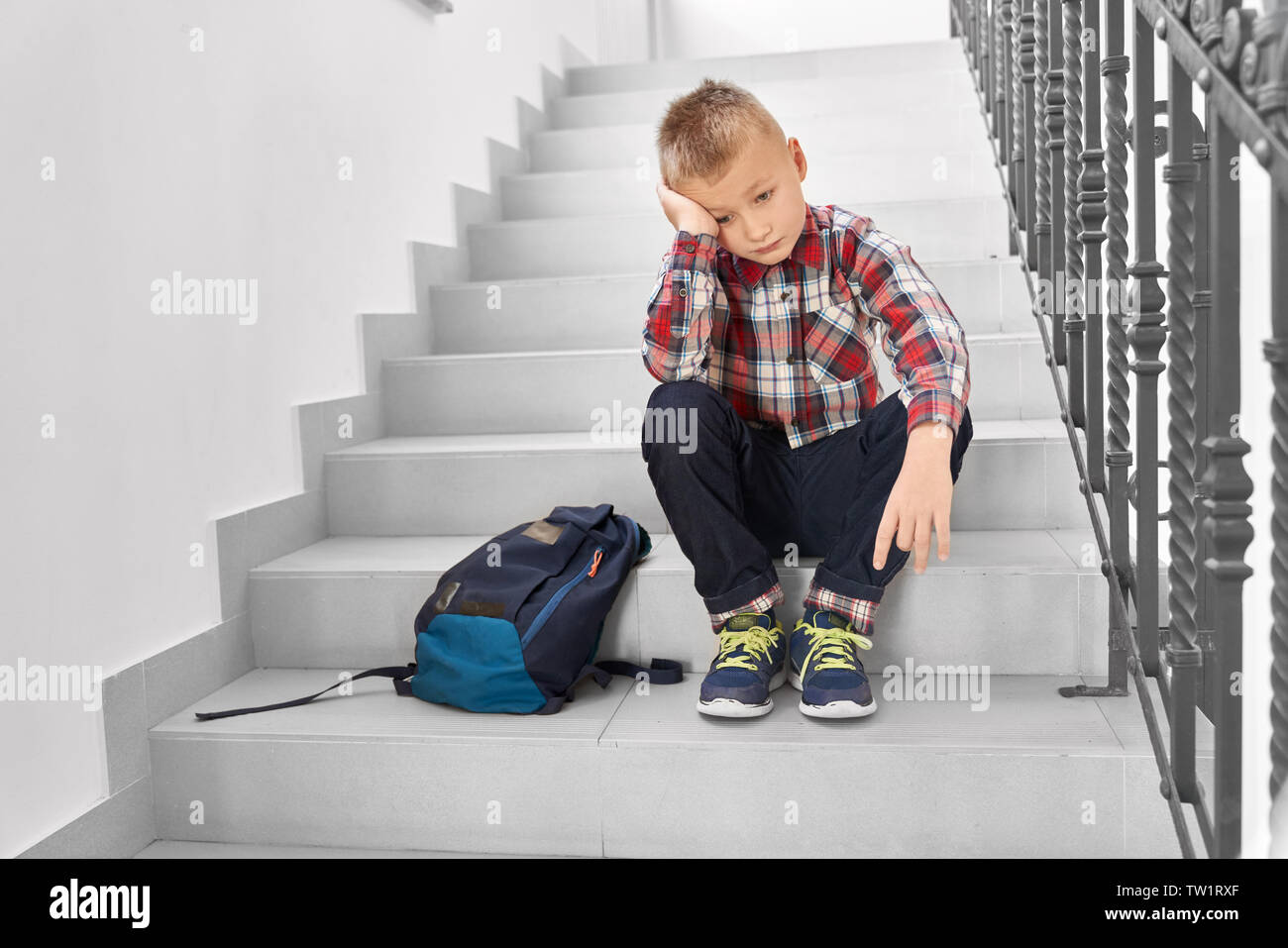 Front view of sad blond boy sitting on stairs in corridor of school and ...