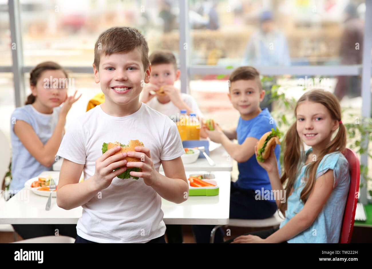 Cute boy eating hamburger and children sitting at table in school ...