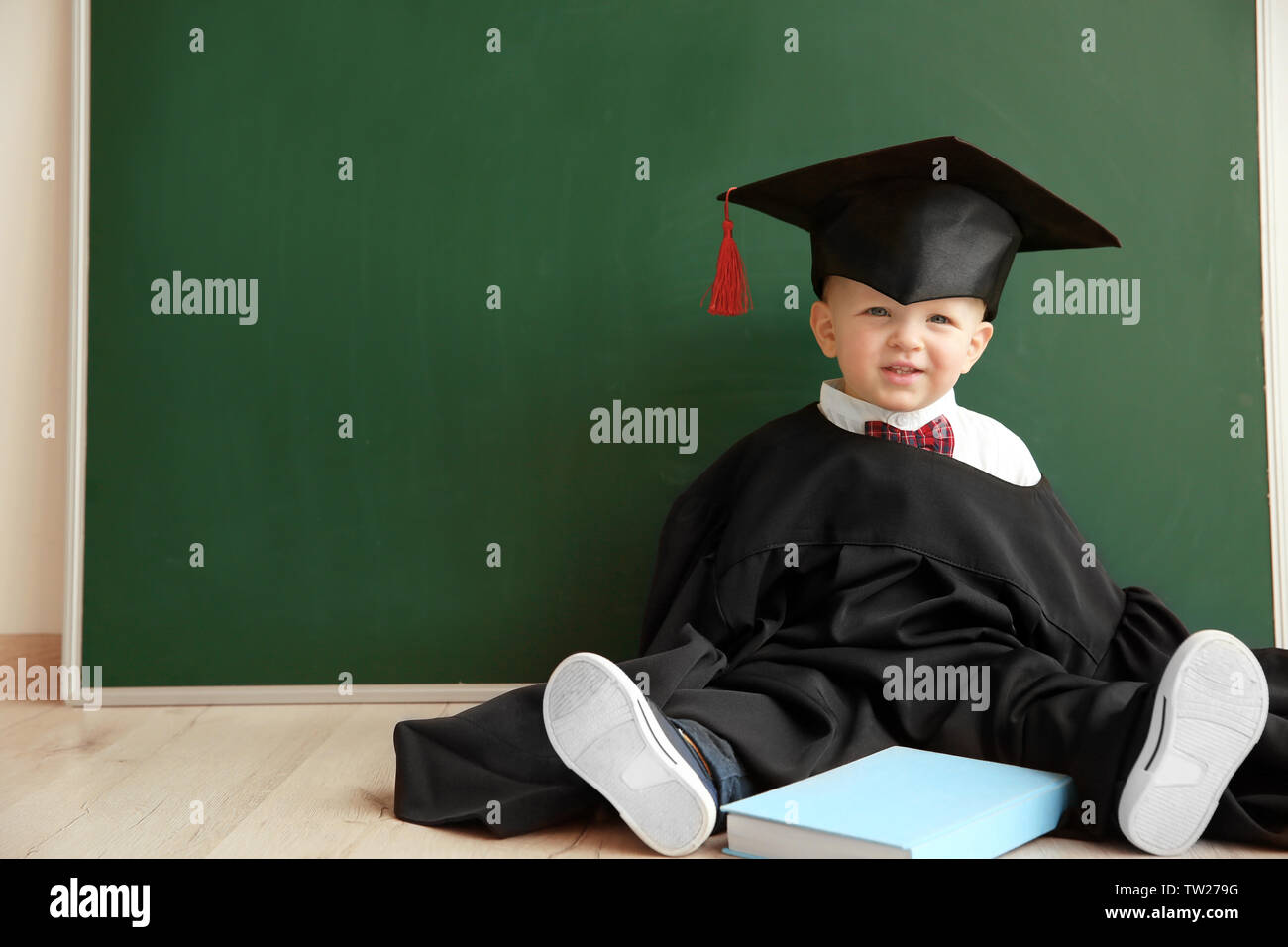 Cute little boy with book, magister hat and gown sitting on floor near ...
