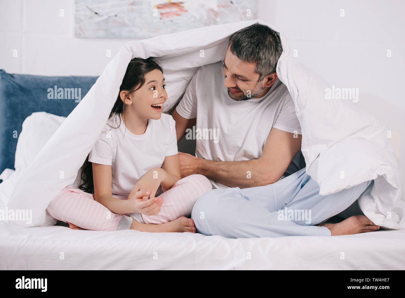 cheerful father sitting with happy daughter and holding blanket on bed ...